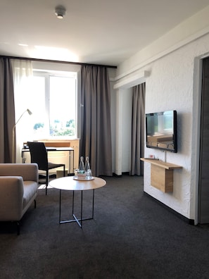 Quarto de hotel com sofá, uma mesinha redonda com garrafas, escrivaninha com cadeira, televisão na parede e uma grande janela com cortinas.