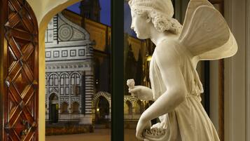 Uma estátua de anjo com asas, segurando uma flor, em frente a um edifício com janelas em arco e uma torre do relógio.