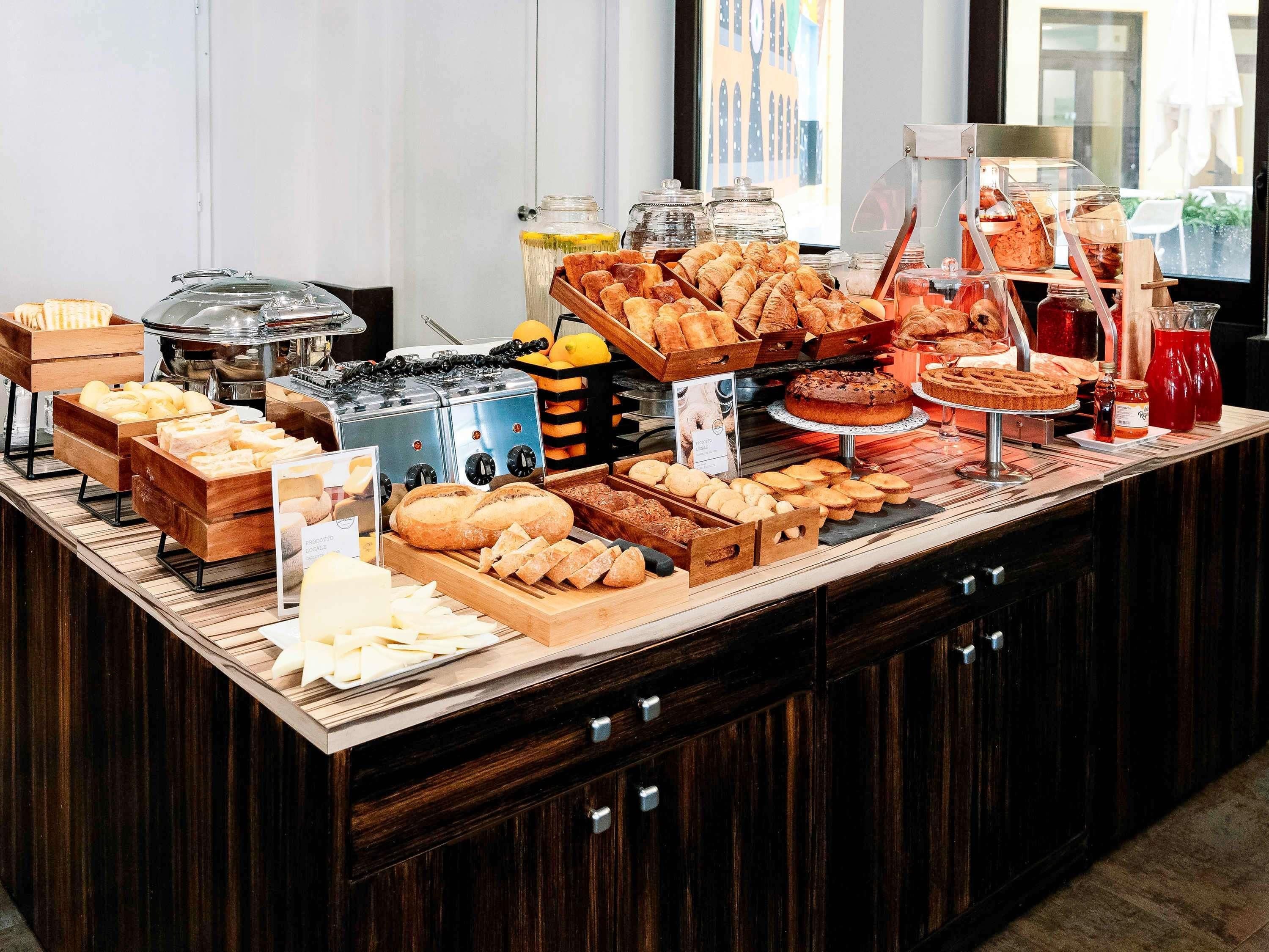 Um buffet com diversos produtos de panificação, incluindo pão, doces e bolos, exposto em um balcão de madeira com vista para uma janela ao fundo.