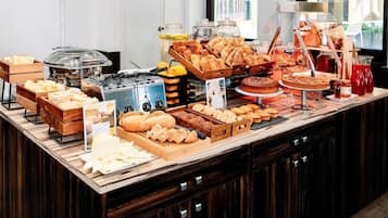 Um buffet com diversos produtos de panificação, incluindo pão, doces e bolos, exposto em um balcão de madeira com vista para uma janela ao fundo.