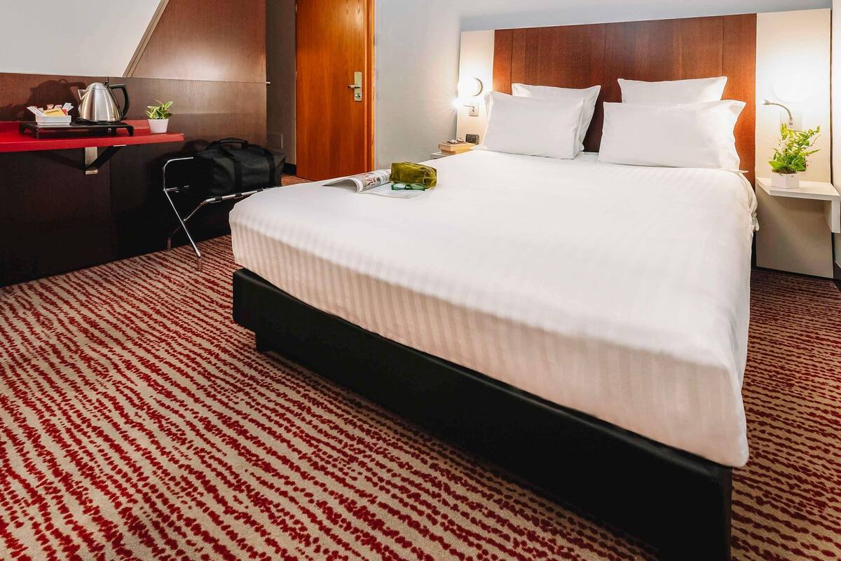 Quarto de hotel com uma cama grande, uma escrivaninha com chaleira, uma pequena planta e um tapete vermelho.