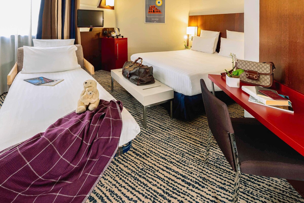 Quarto de hotel com duas camas, uma escrivaninha, uma cadeira, uma televisão, um armário vermelho e um tapete estampado.