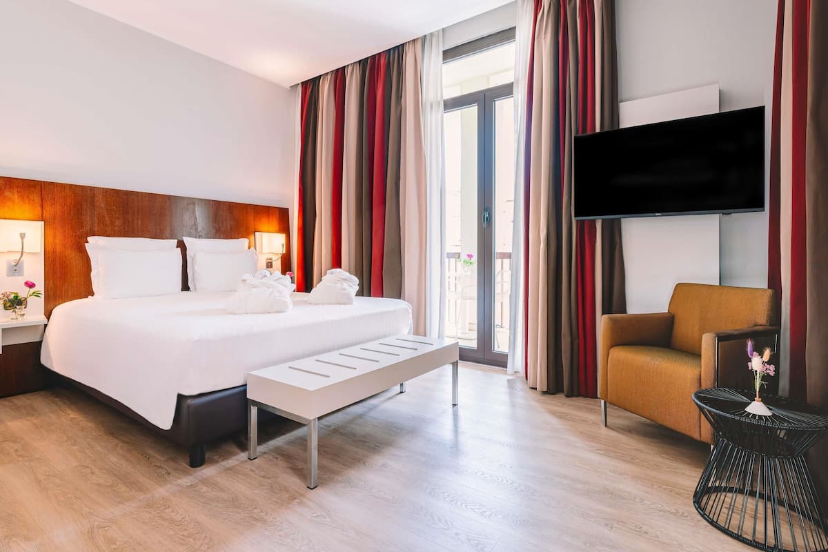 Quarto de hotel com cama, mesa de cabeceira, luminária, televisão, poltrona e uma pequena mesa com flores.