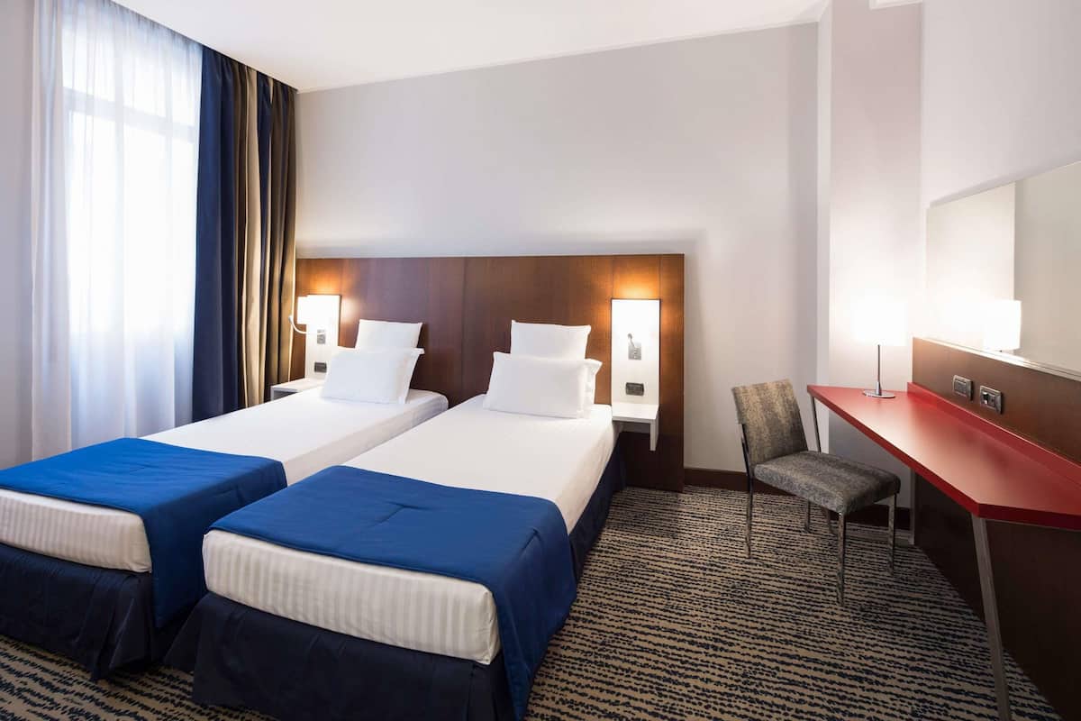 Quarto de hotel com duas camas, uma escrivaninha, uma cadeira e uma janela grande com cortinas.