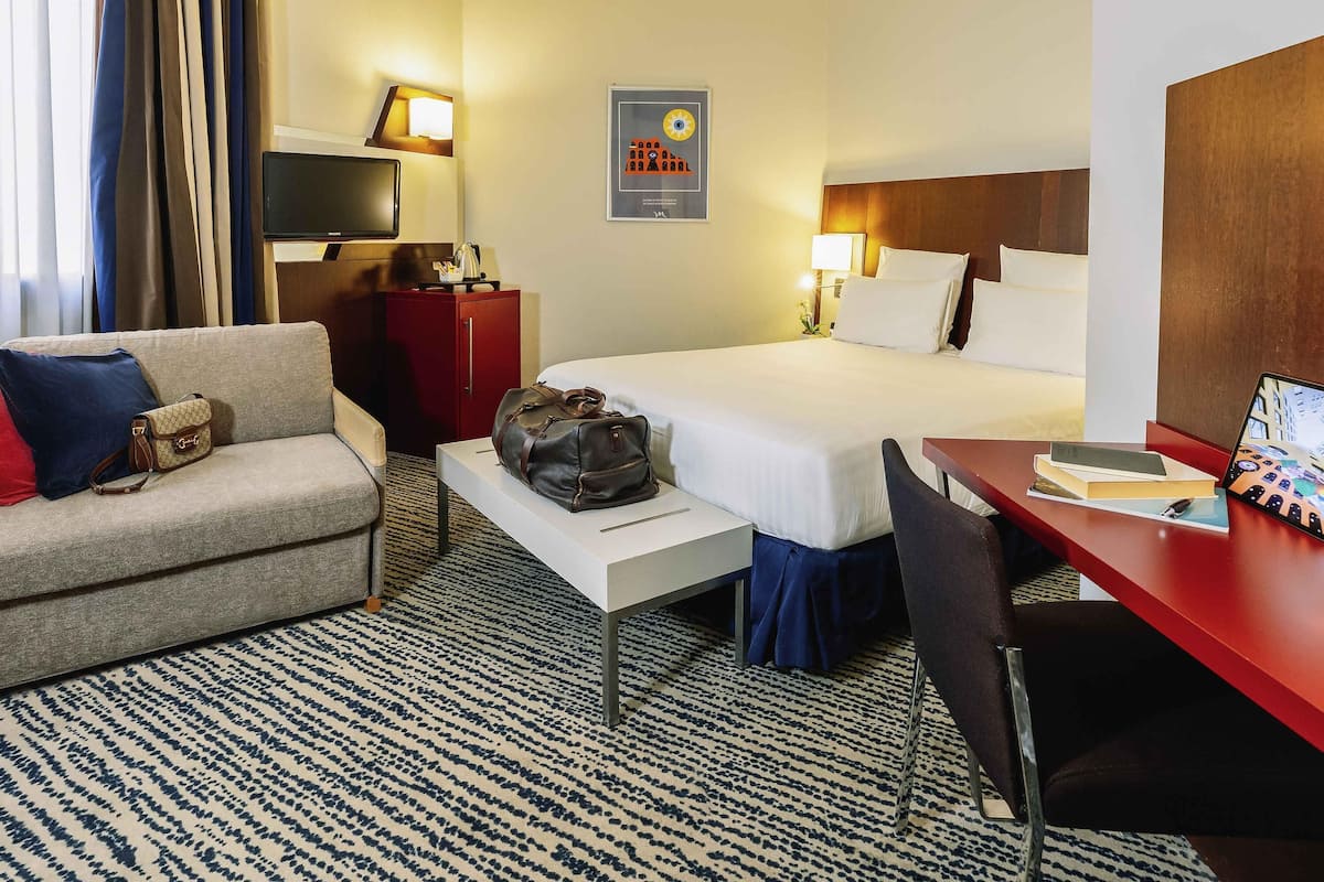 Quarto de hotel com cama, escrivaninha, cadeira, sofá, armário vermelho e televisão na parede.