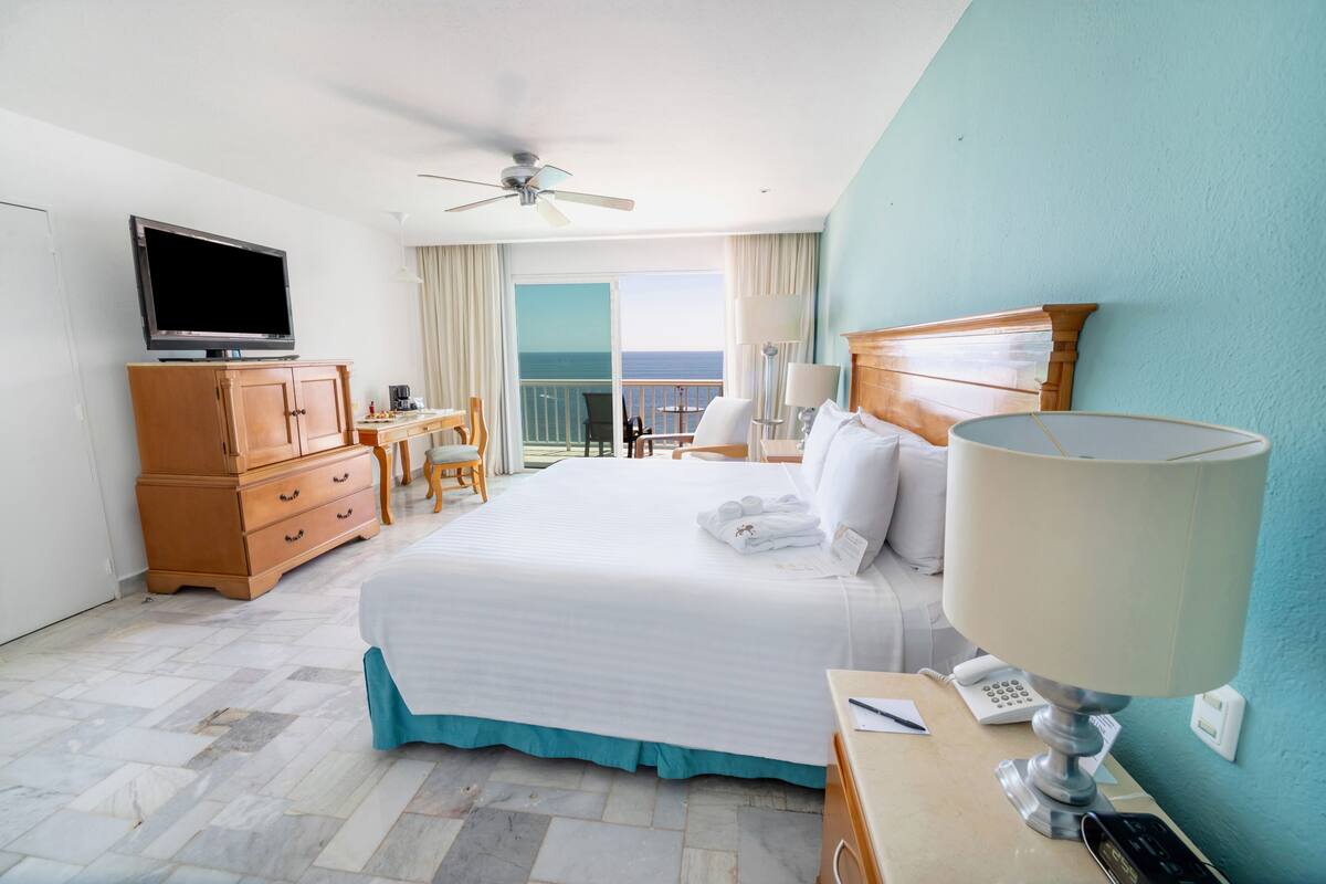 Quarto de hotel com cama, televisão, escrivaninha com luminária, telefone e vista para o oceano.