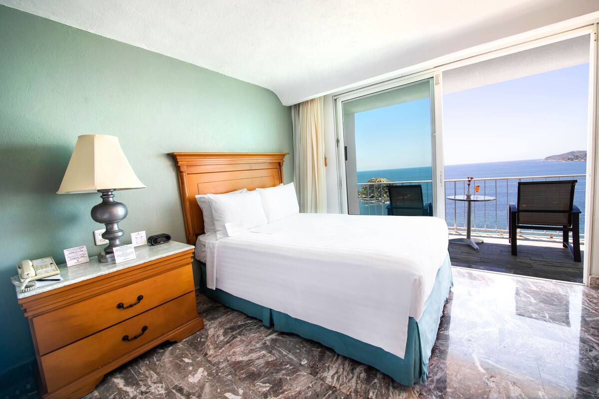 Quarto de hotel com cama, mesa de cabeceira, luminária, escrivaninha com telefone e vista para o oceano através de uma varanda.
