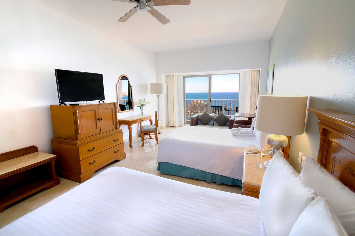 Quarto de hotel com duas camas, uma escrivaninha, cômoda, televisão e varanda com vista para o mar.