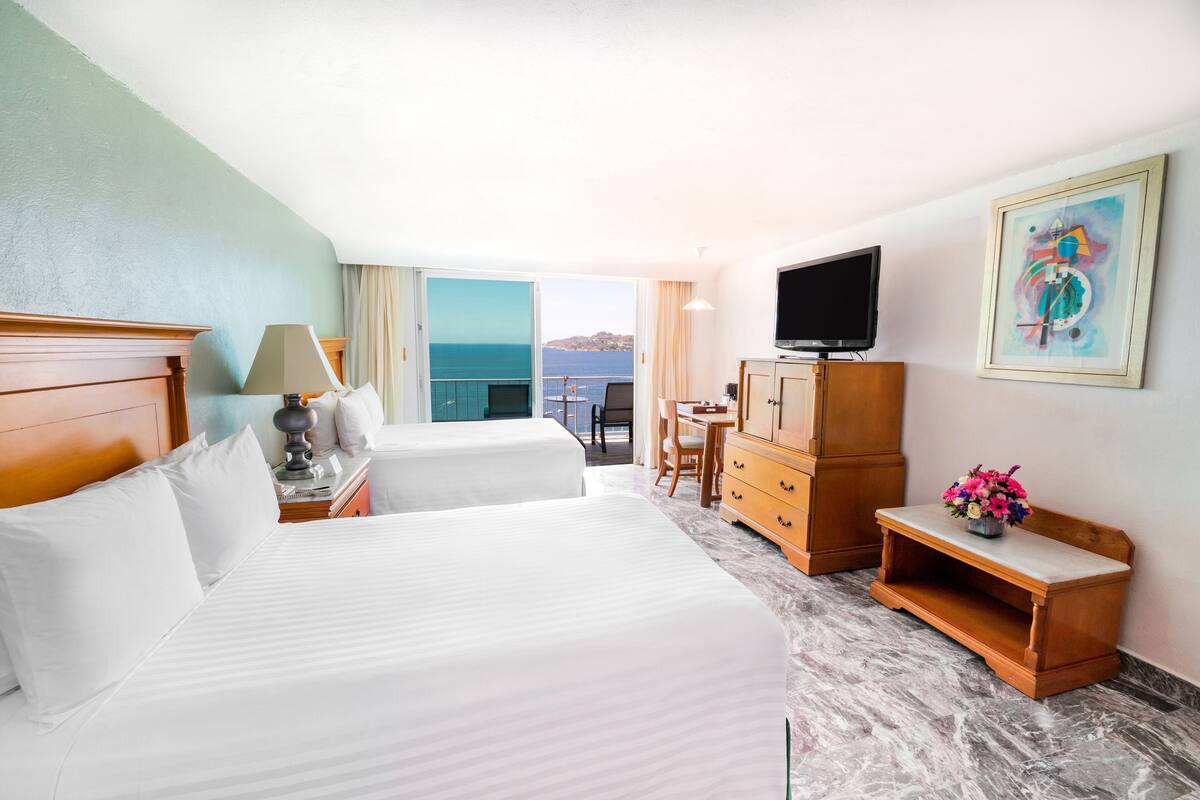 Quarto de hotel com duas camas, televisão, escrivaninha e vista para o mar.