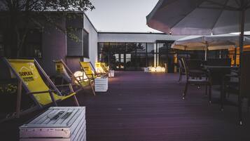 Um deck externo com assentos e guarda-sóis, um edifício com grandes janelas e uma placa com 'SOPLICA'.