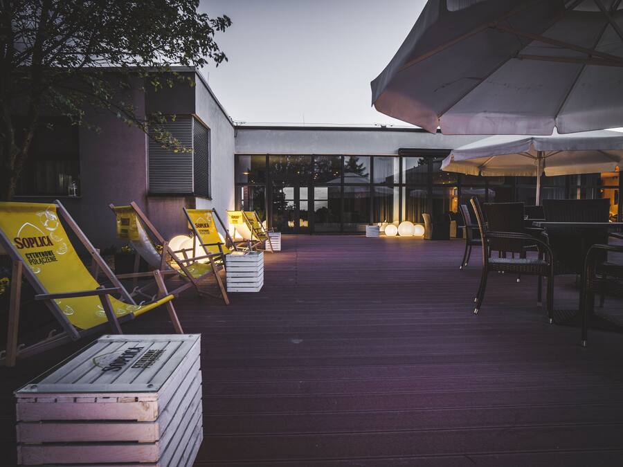 Um deck externo com assentos e guarda-sóis, um edifício com grandes janelas e uma placa com 'SOPLICA'.