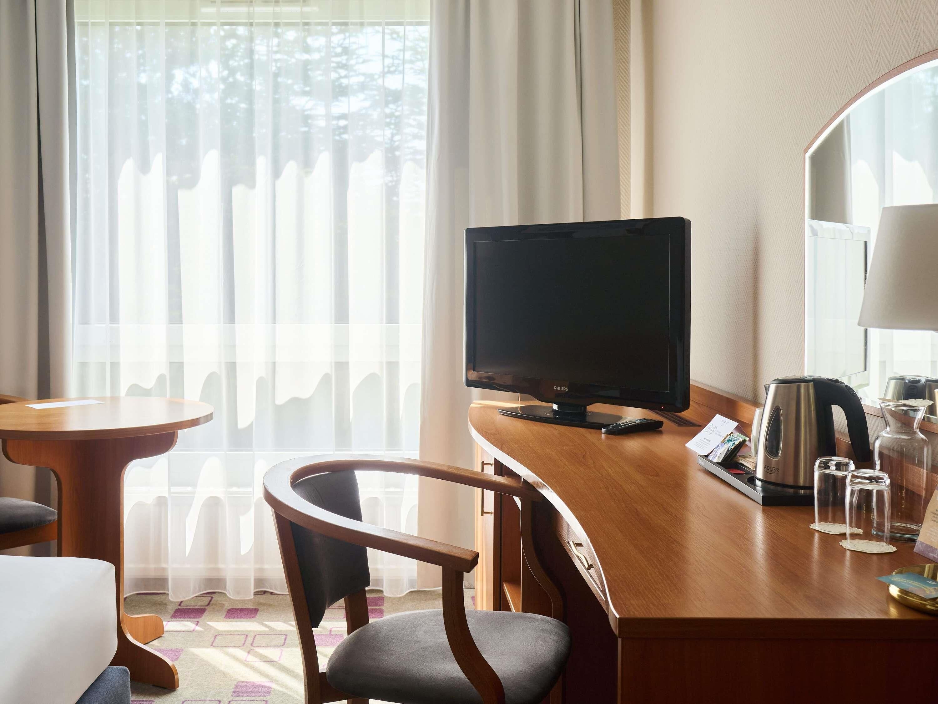 Quarto de hotel com escrivaninha de madeira, televisão, cadeira, cafeteira e janela com cortinas finas.