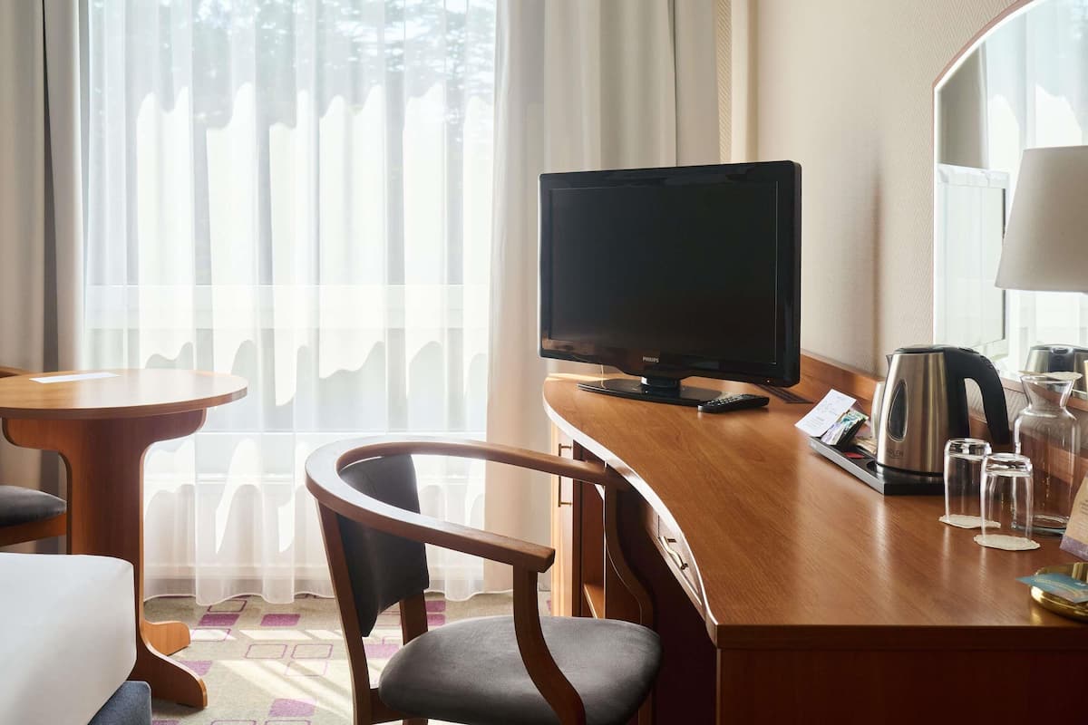 Quarto de hotel com escrivaninha de madeira, televisão, cadeira, cafeteira e janela com cortinas finas.