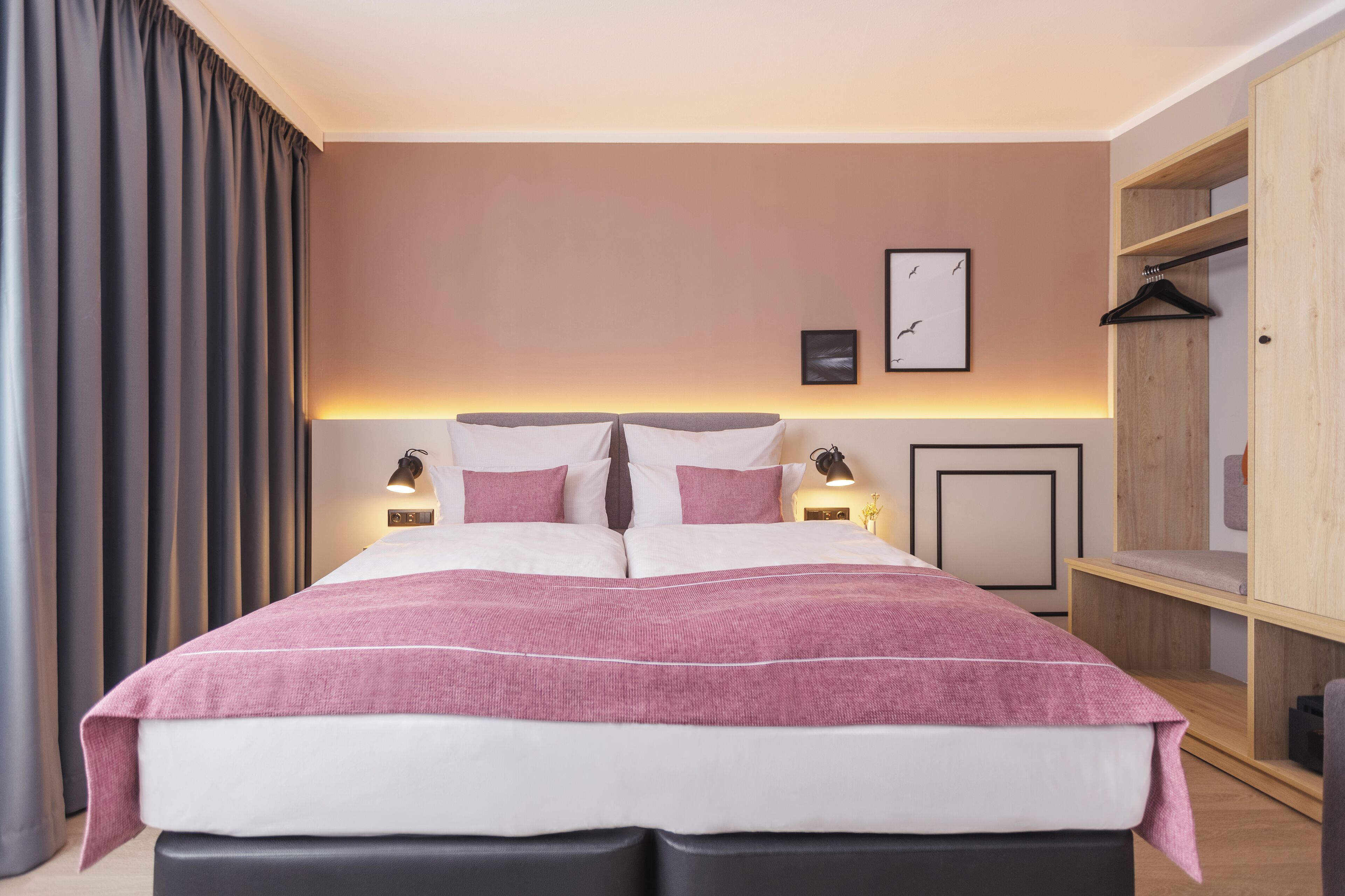Uma cama bem arrumada com um cobertor rosa, duas luminárias laterais e um guarda-roupa de madeira com cabideiro.