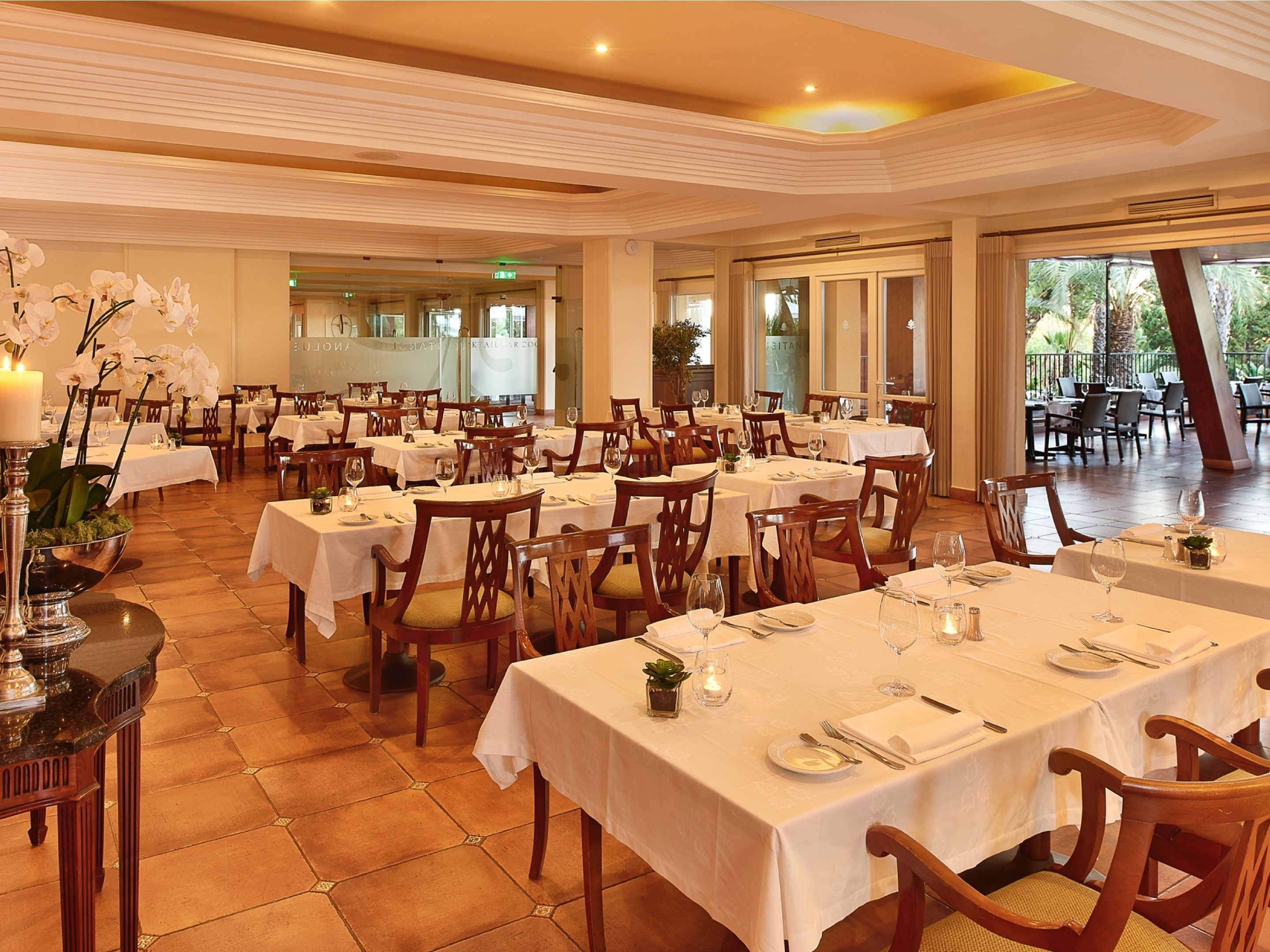 Uma sala de jantar ampla, com várias mesas e cadeiras, um lustre e vista para o exterior.