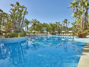 Piscina com bordas azulejadas, rodeada por espreguiçadeiras e guarda-sóis, com palmeiras ao fundo.