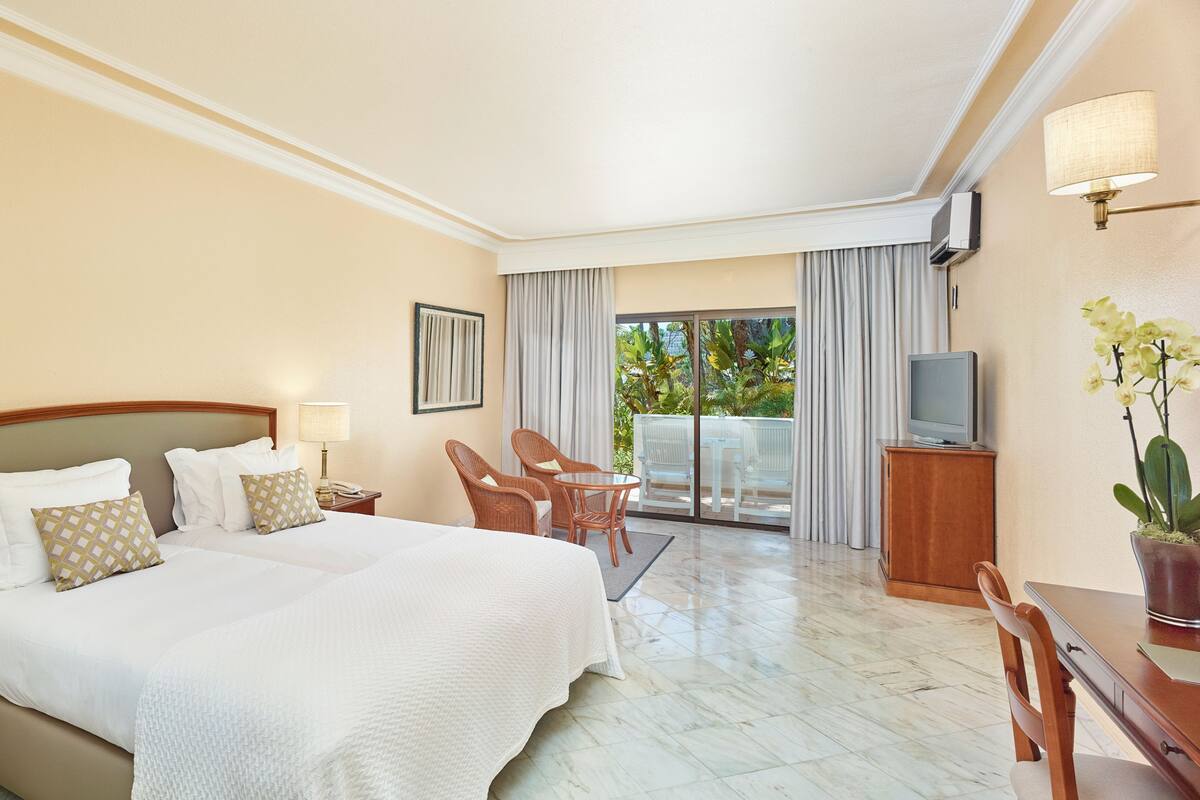 Quarto de hotel com cama grande, escrivaninha com cadeira, televisão e varanda com cortinas.
