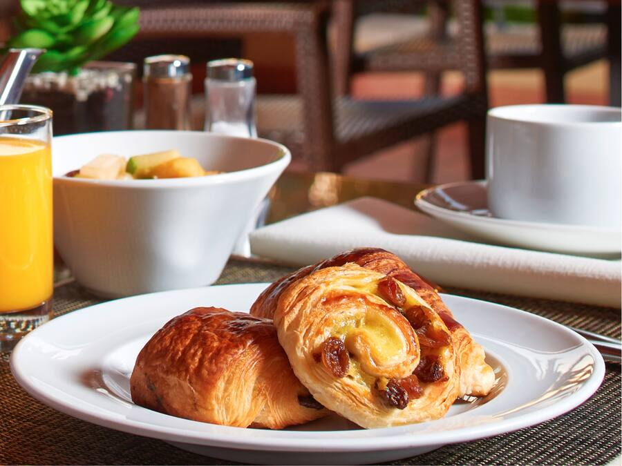 Uma mesa com croissants, uma tigela de fruta e um copo de suco de laranja para o café da manhã.