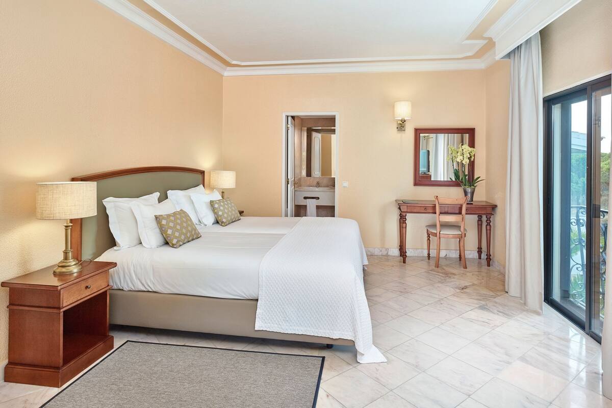 Quarto de hotel com uma cama grande, mesas de cabeceira, escrivaninha, cadeira, espelho e vista para o exterior.