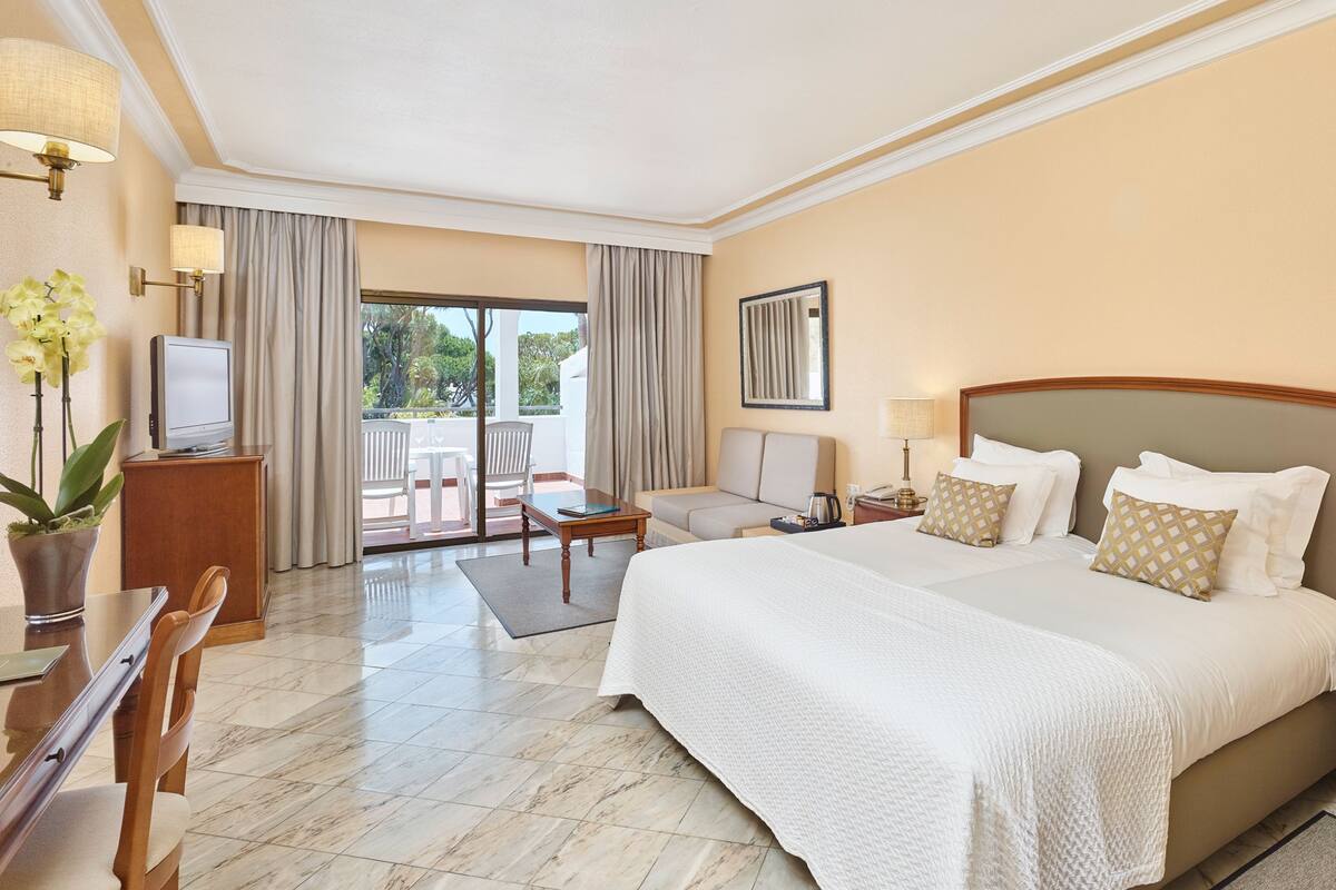Quarto de hotel com uma cama grande, escrivaninha, cadeira, televisão e varanda com mesa e cadeiras.