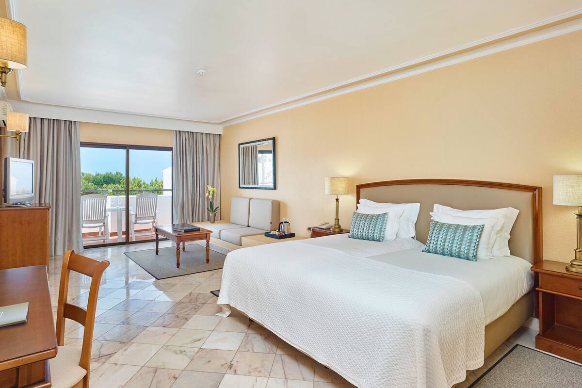 Quarto de hotel com uma cama grande, uma escrivaninha, uma cadeira, um sofá e uma varanda com mesa e cadeiras.