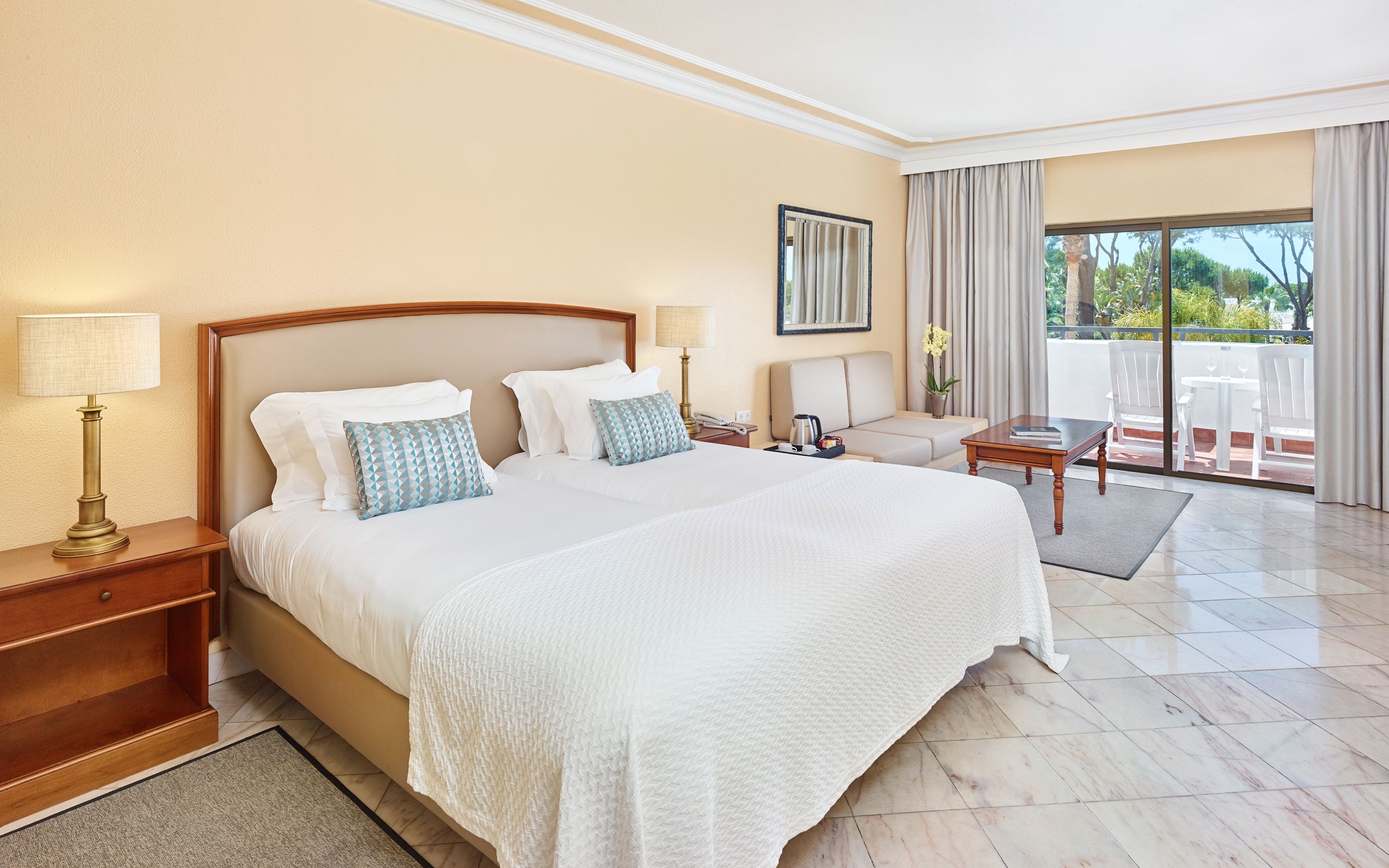 Quarto de hotel com uma cama grande, uma mesa de cabeceira com luminária, uma mesinha com um livro, uma cadeira e vista para o exterior por uma porta deslizante.