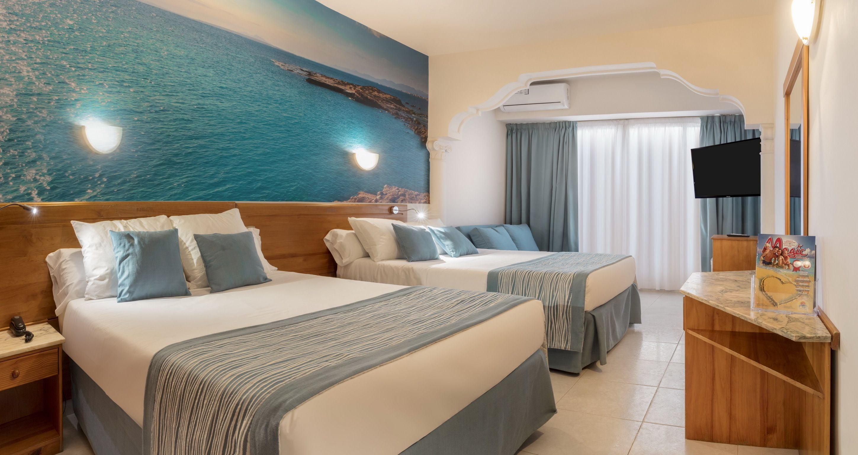 Quarto de hotel com duas camas, um mural grande com vista para o oceano, uma TV de tela plana e uma mesa de cabeceira de madeira.