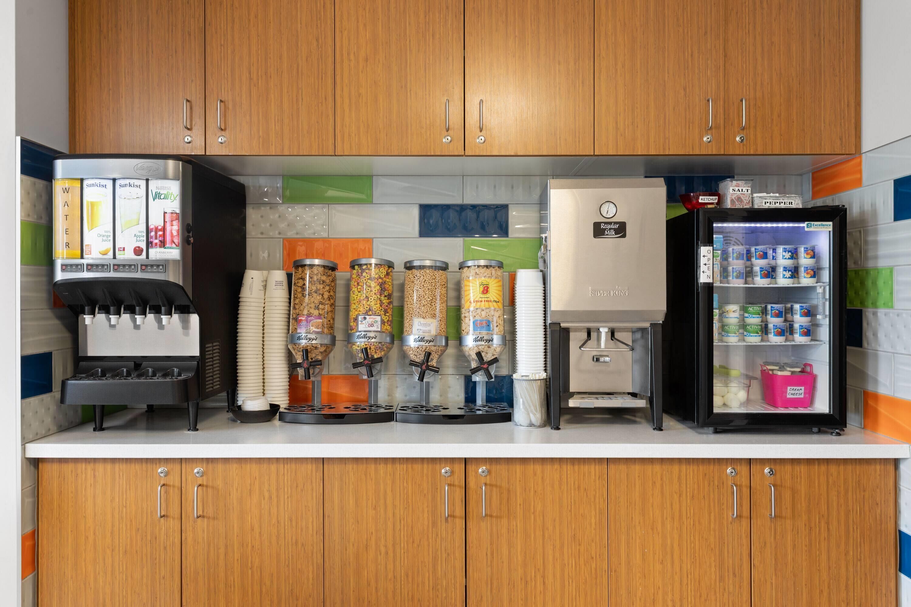 Uma área de cozinha com bebedouro, cafeteira, dispensadores de cereais, frigorífico e mini-frigorífico.