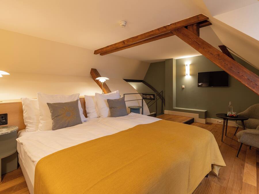 Quarto com cama, mesa de cabeceira, luminária, poltrona e televisão na parede.