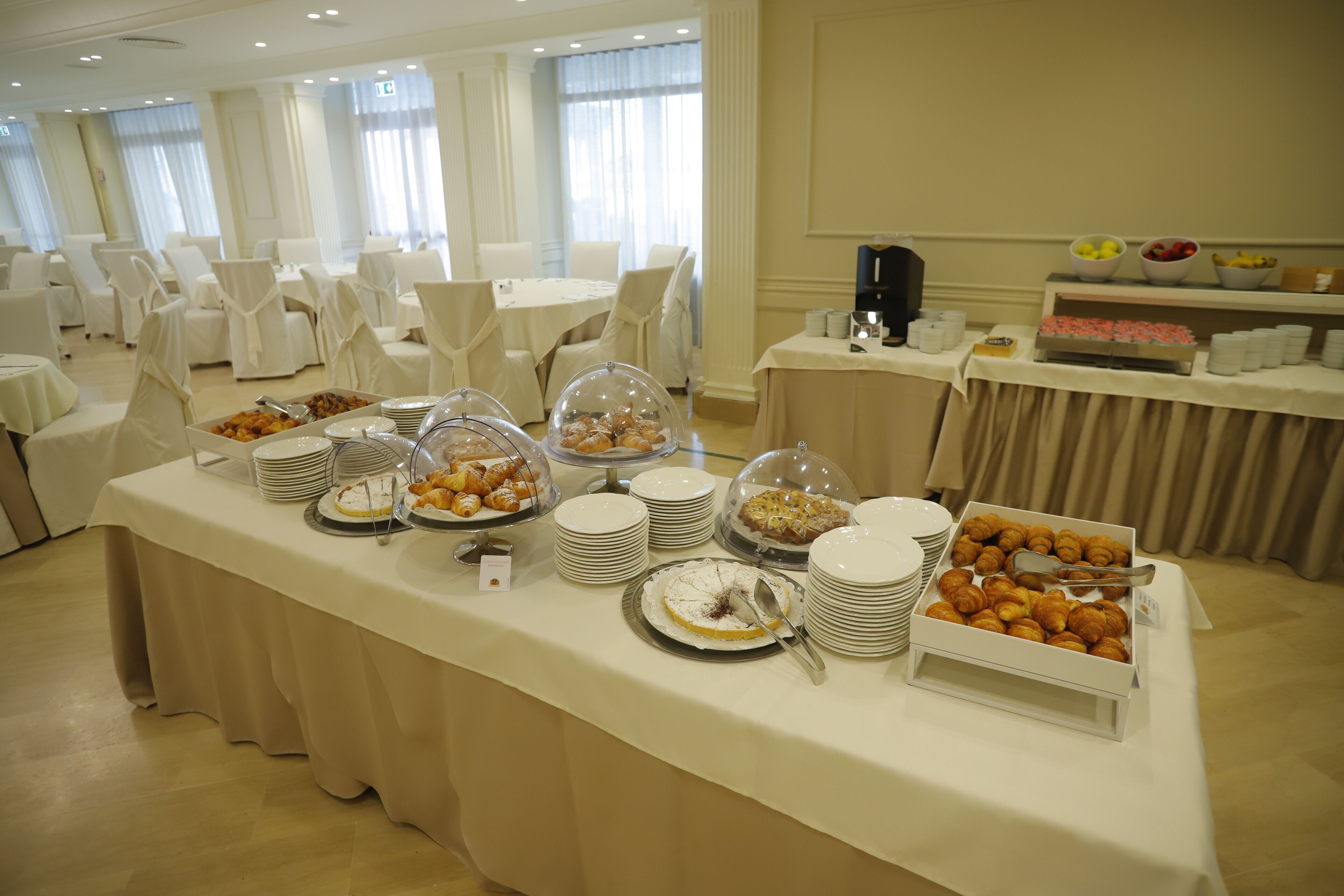 Disposição de buffet com diversos alimentos e área de estar com cadeiras e mesas brancas.