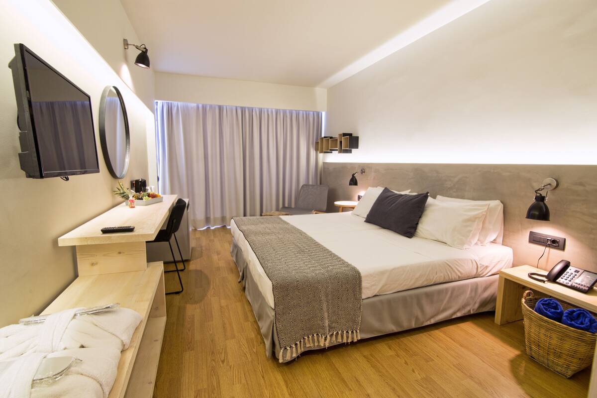 Quarto de hotel moderno com uma cama grande, uma escrivaninha com computador, uma TV e um banheiro com chuveiro e vaso sanitário.