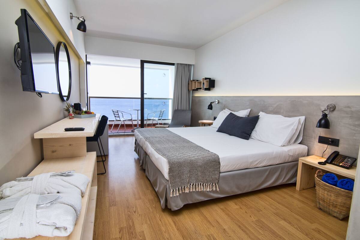 Quarto de hotel moderno com cama, escrivaninha e cadeira, uma janela grande com vista e uma varanda.