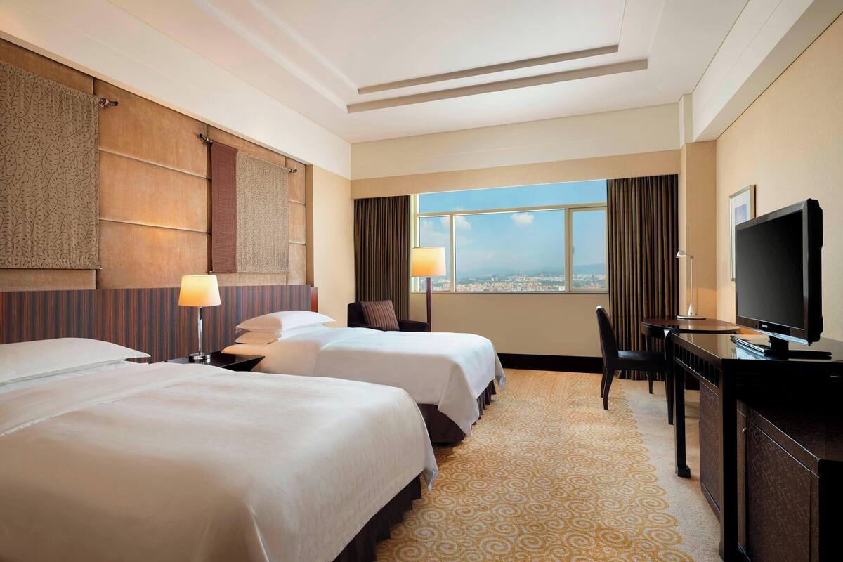Quarto de hotel com duas camas, uma escrivaninha, televisão e vista para a cidade.