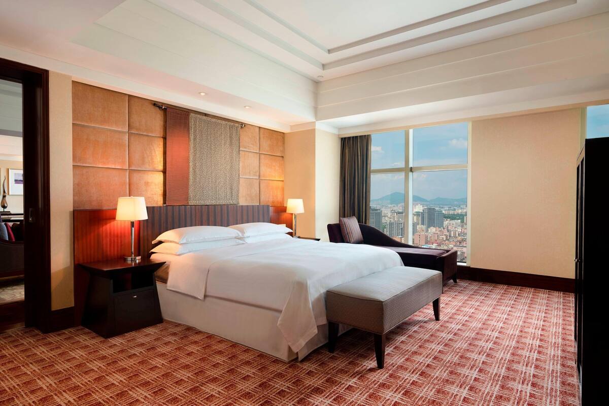 Quarto de hotel com uma cama grande, duas mesas de cabeceira com abajures, uma cadeira e vista para a cidade através de uma janela ampla.