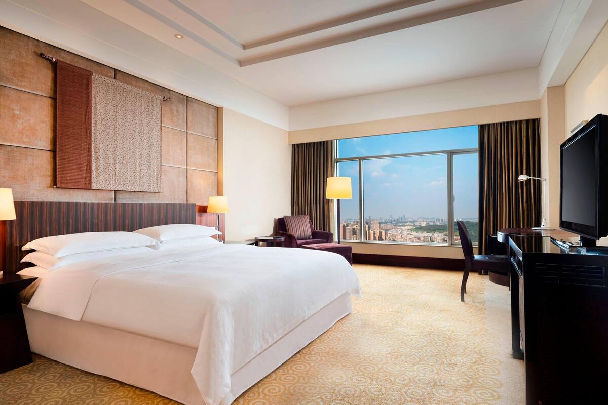 Quarto de hotel com uma cama grande, uma escrivaninha com televisão, uma cadeira e vista para a cidade através de amplas janelas.