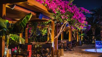 Uma área externa com mesas e cadeiras de madeira, cercada por vibrantes flores de bougainvillea rosa e uma estrutura de pérgola de madeira.