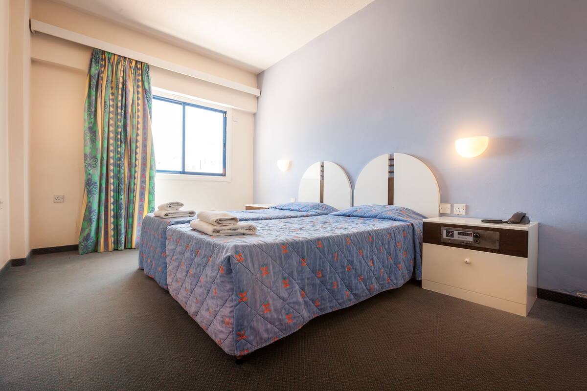 Quarto de hotel com cama, mesa de cabeceira, abajur e janela com cortinas.