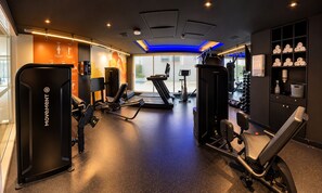 Ginásio com diversos equipamentos de exercício, incluindo esteiras e aparelhos de musculação, e uma parede com suporte para toalhas.