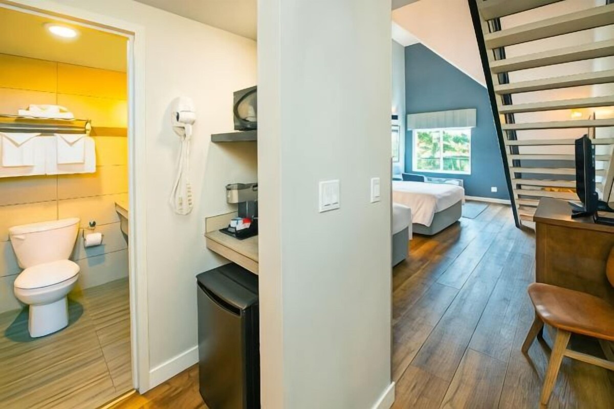 Quarto de hotel compacto com banheiro, uma pequena cozinha, uma cama e uma escada que leva a um andar superior.