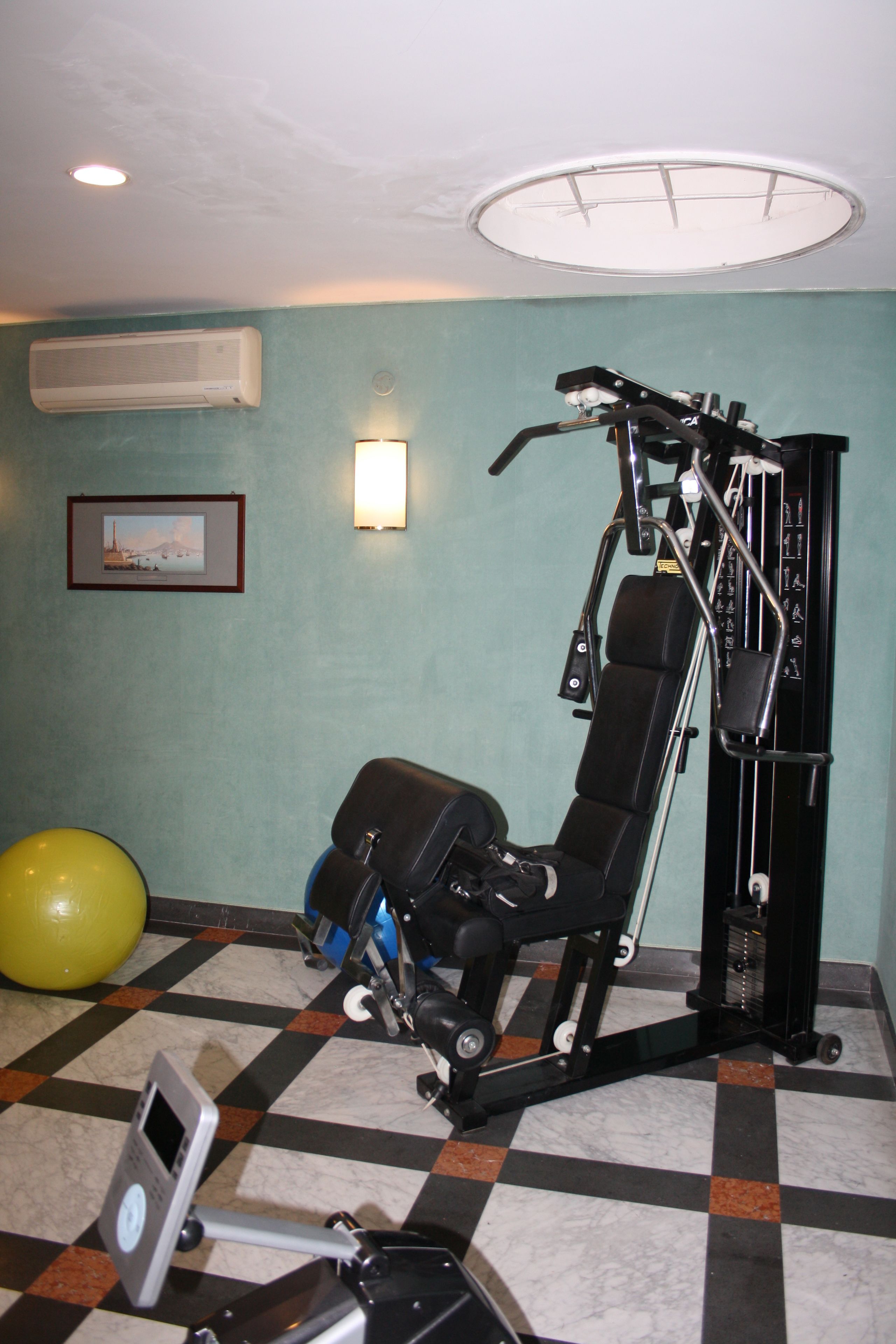 Uma academia com equipamentos de ginástica, uma bola de exercícios amarela e um ar condicionado fixado na parede.