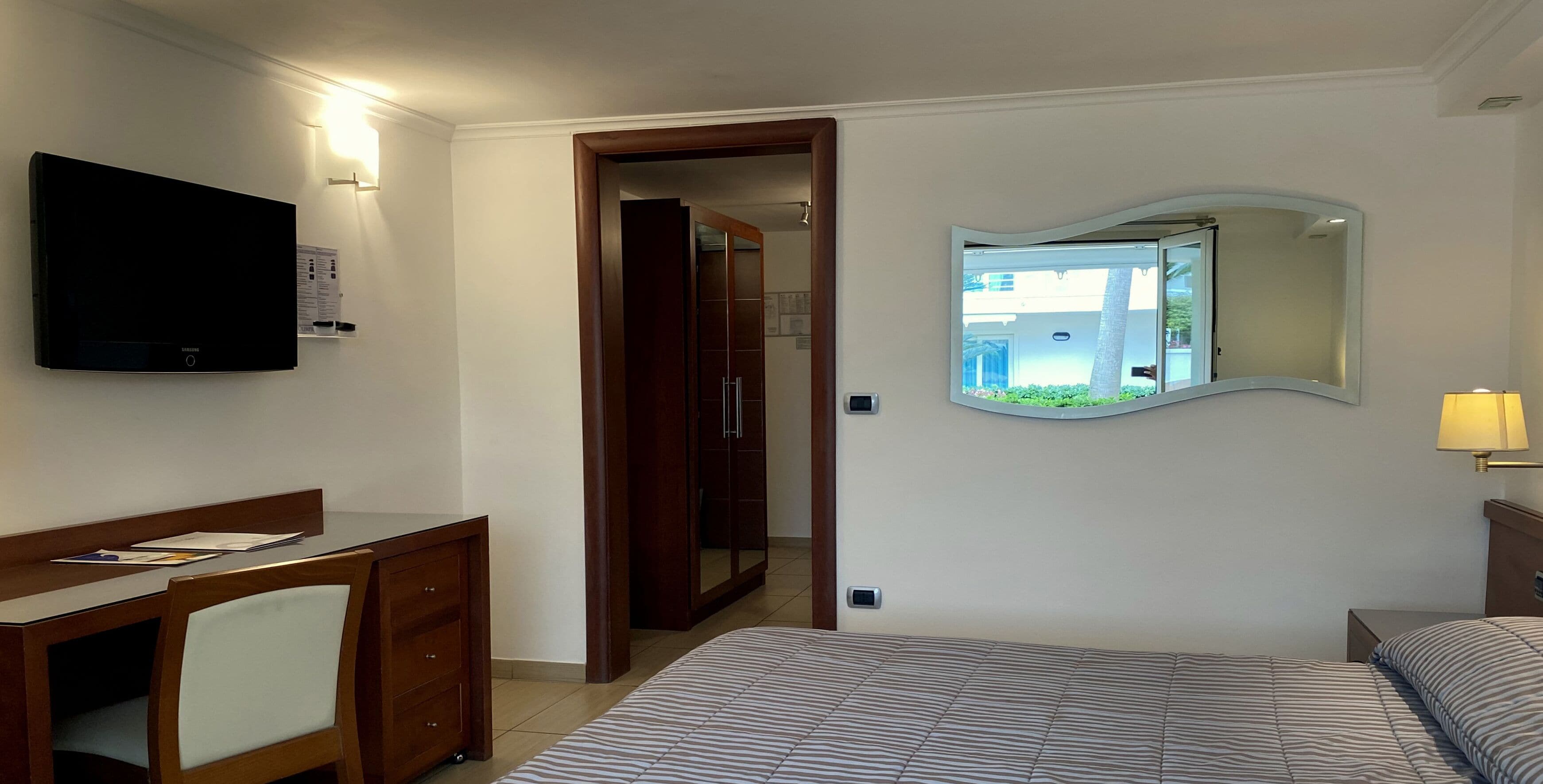 Quarto de hotel com cama, escrivaninha com papéis, televisão, guarda-roupa e janela com vista para o exterior.