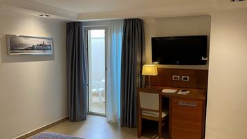 Quarto de hotel com uma cama, uma mesa com cadeira, uma televisão, um quadro na parede e uma porta de correr para uma varanda.
