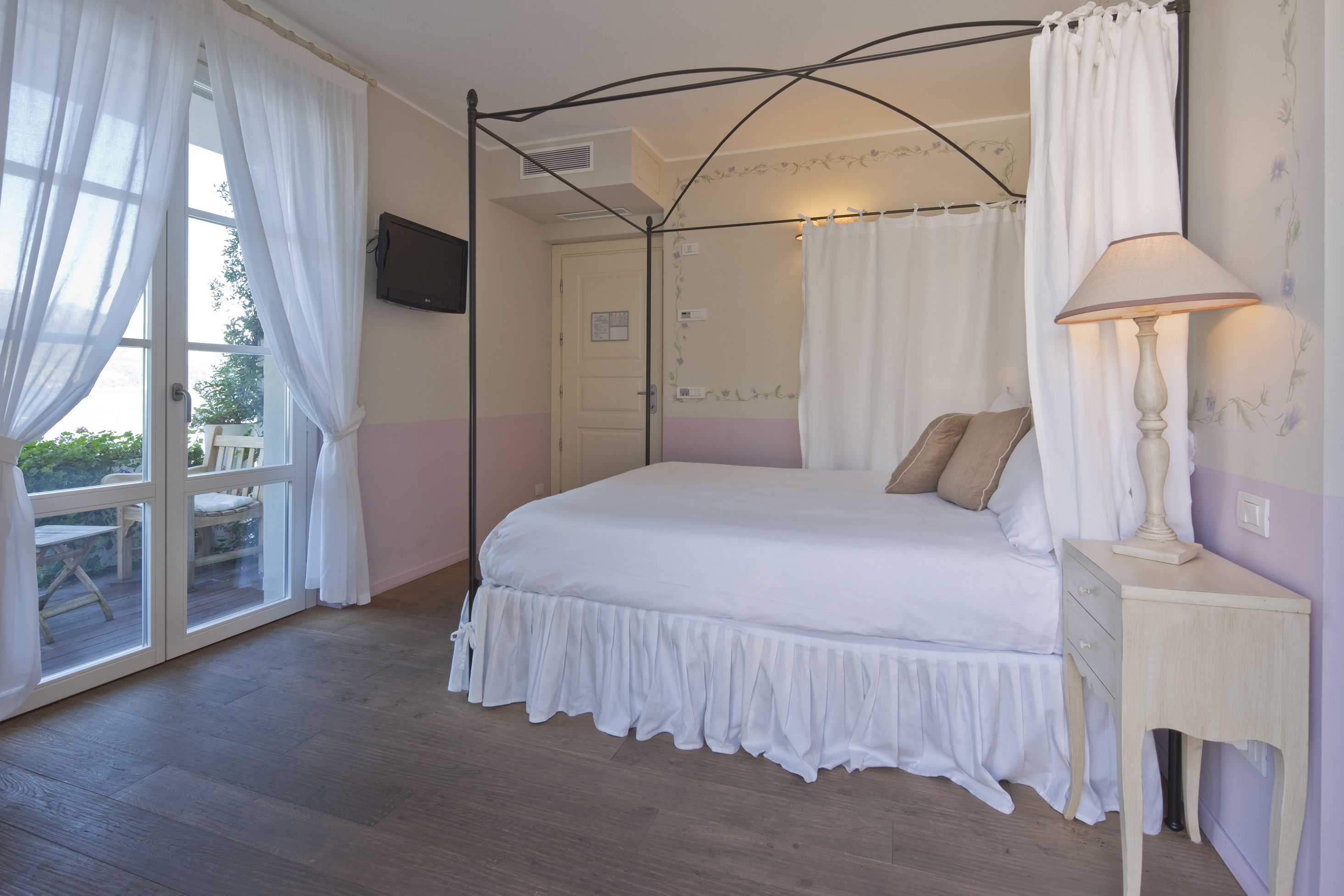 Quarto com cama de dossel, mesa de cabeceira com abajur e vista através de uma janela grande com cortinas.