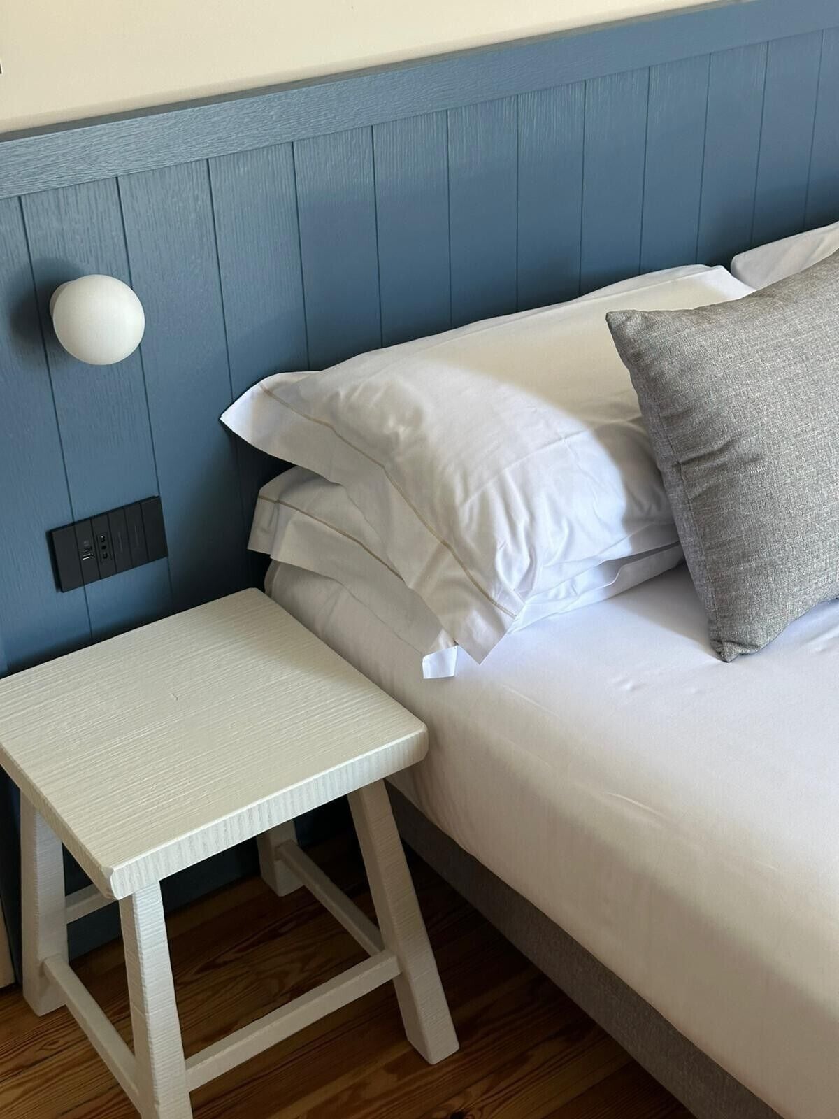 Uma cama com roupa de cama branca, um travesseiro cinza e uma mesa de cabeceira branca.