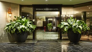 Entrada do Hotel Brisbane Riverview, com chão de mármore, grandes vasos de plantas e um candelabro visível no interior.