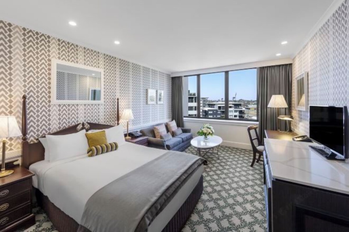 Quarto de hotel com cama, sofá, televisão e uma grande janela com vista para a cidade.