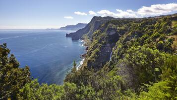 Paisagem costeira com vista clara para o mar, vegetação exuberante e montanhas ao fundo.