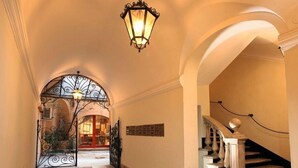 Um amplo salão com um portão de metal ornamentado, uma escadaria com balaústres e um luminário fixado na parede.