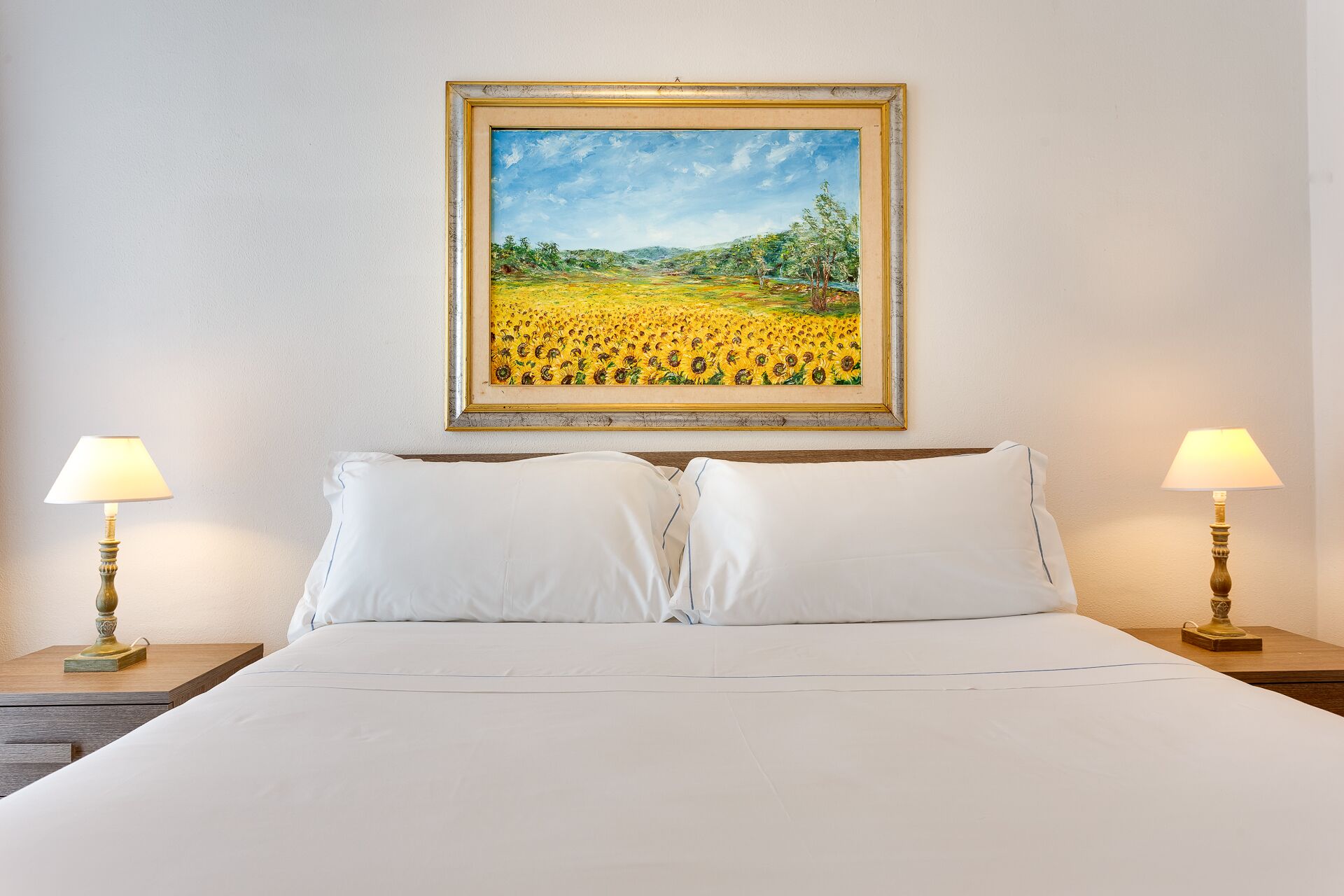 Quarto de hotel com uma cama, duas luminárias de cabeceira e um quadro de um campo.