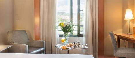 Quarto de hotel com uma cama, duas poltronas, uma mesa pequena com itens de café da manhã, uma janela com cortinas e um abajur.